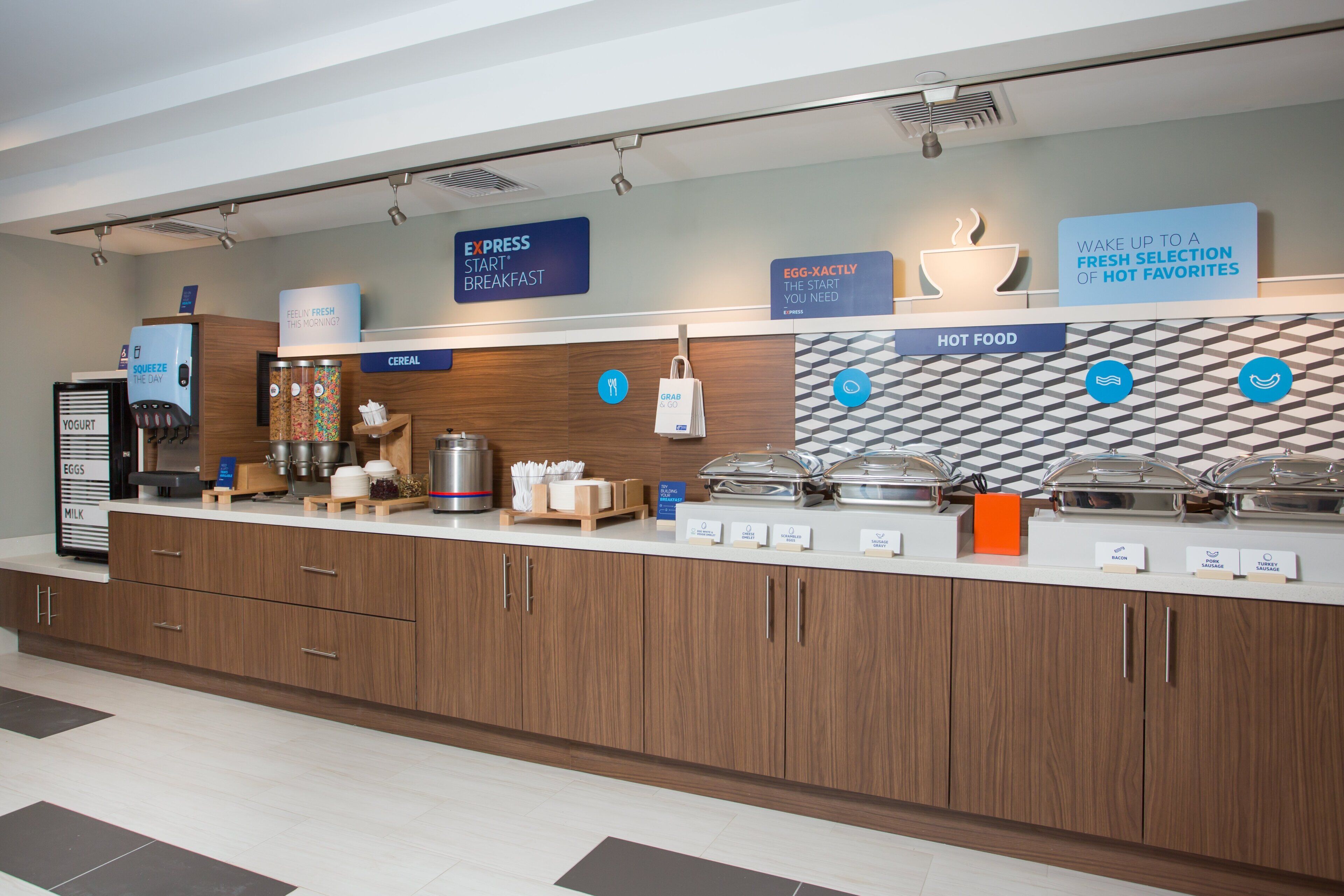 Espaço de café da manhã de hotel com estação de autoatendimento, incluindo iogurte, ovos, leite, cereais e opções de comida quente como salsicha e salsicha de peru.