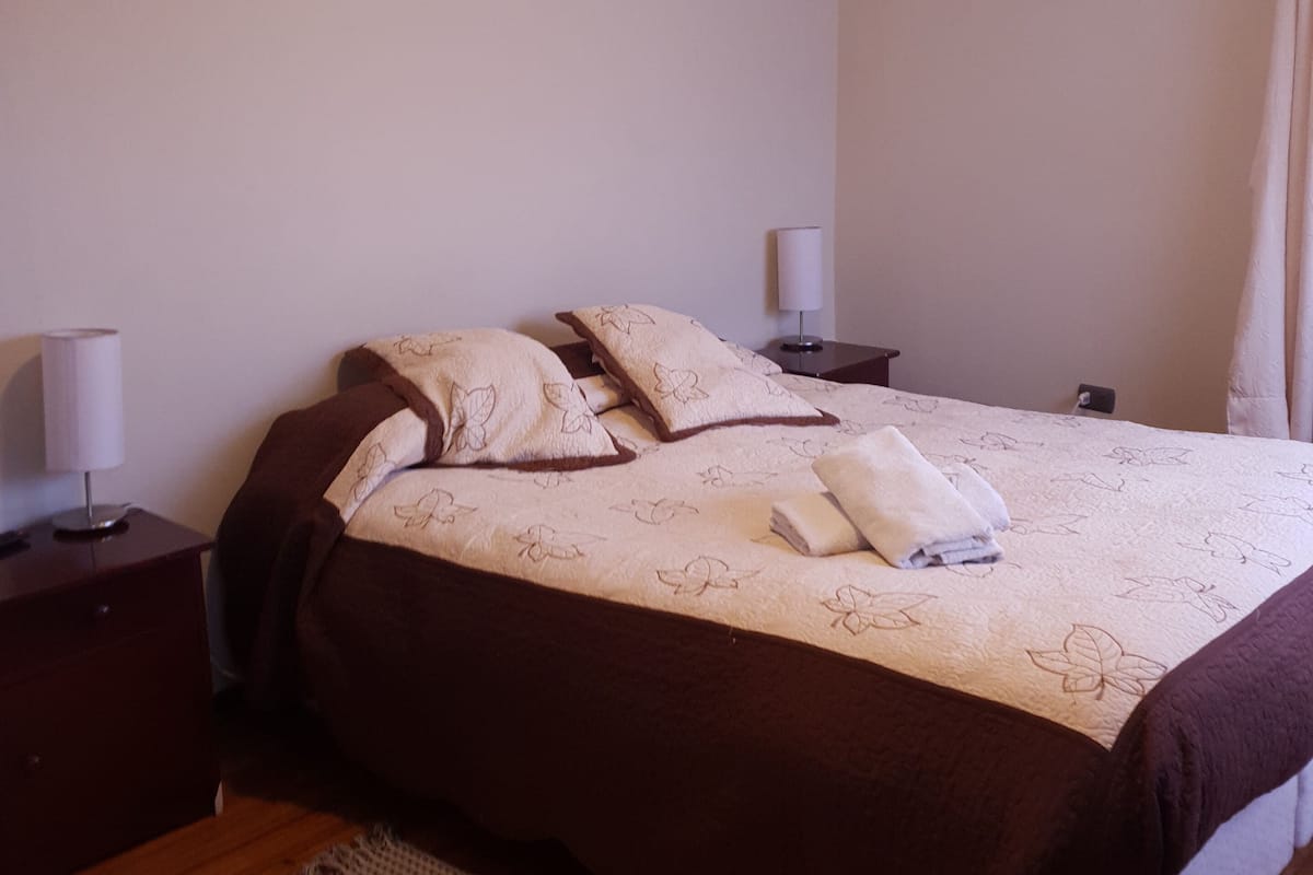 Uma cama bem arrumada com colcha florida, duas mesas de cabeceira com abajures, uma moldura pequena na parede e piso de madeira.