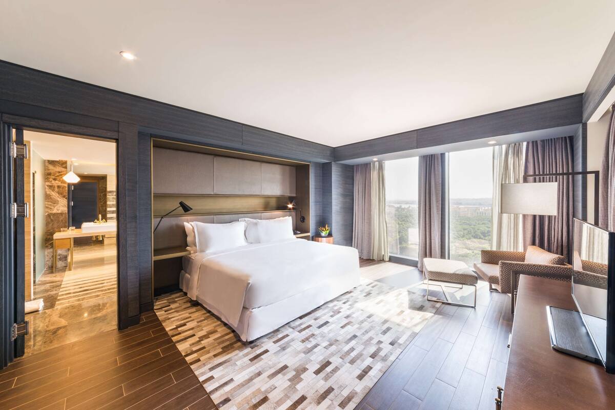 Quarto de hotel moderno com uma cama grande, televisão, escrivaninha e vista para a cidade.
