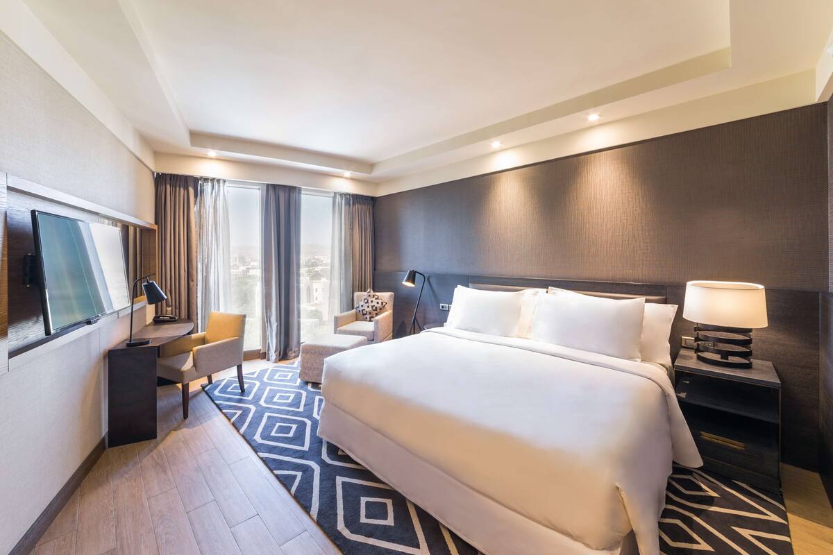 Quarto de hotel com uma cama grande, uma escrivaninha com cadeira, televisão e vista da cidade através de grandes janelas.
