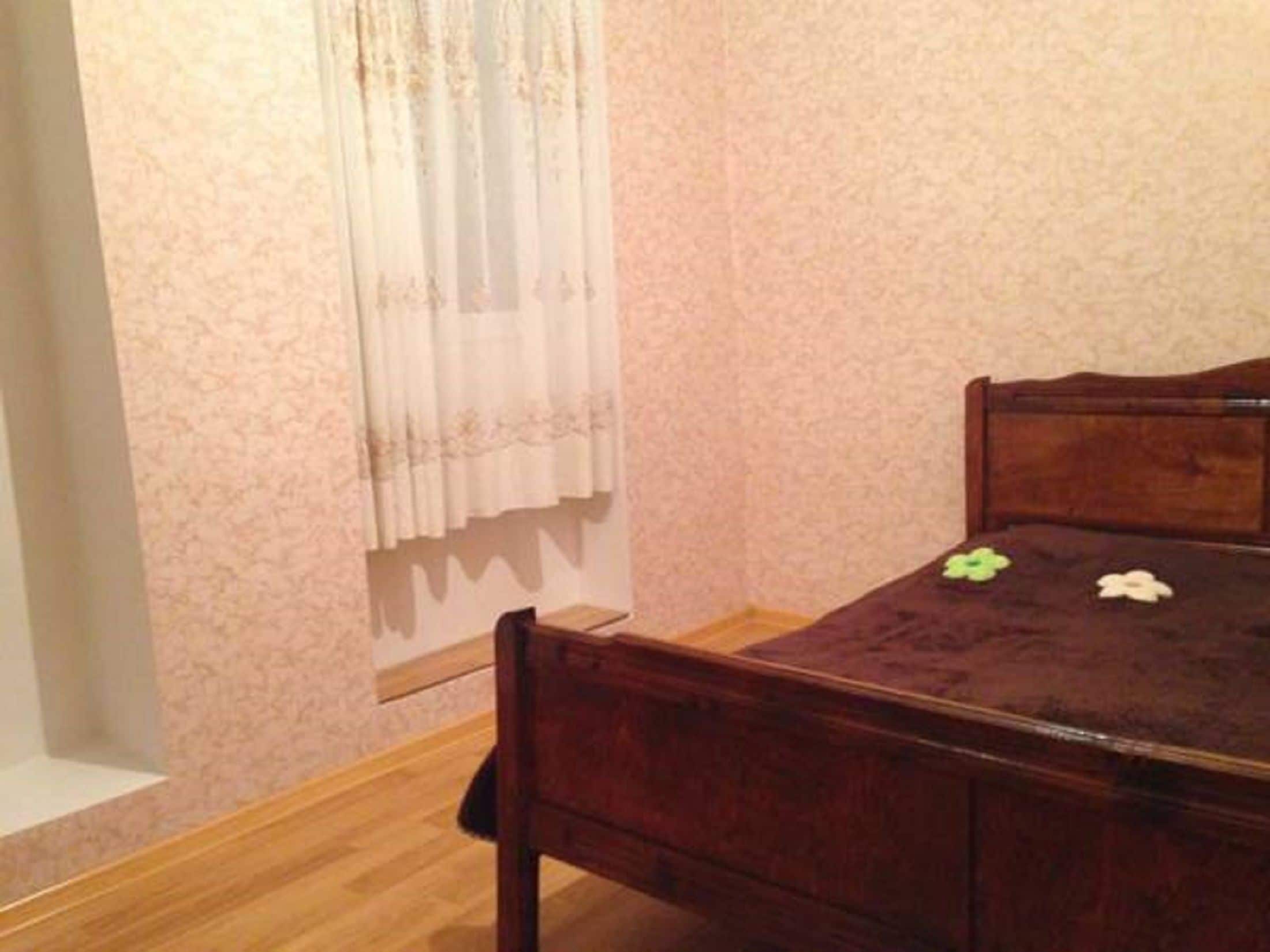 Quarto com cama de madeira, janela com cortinas leves e piso coberto por carpete.