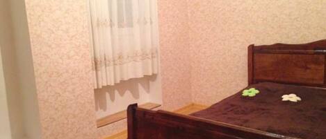 Quarto com cama de madeira, janela com cortinas leves e piso coberto por carpete.