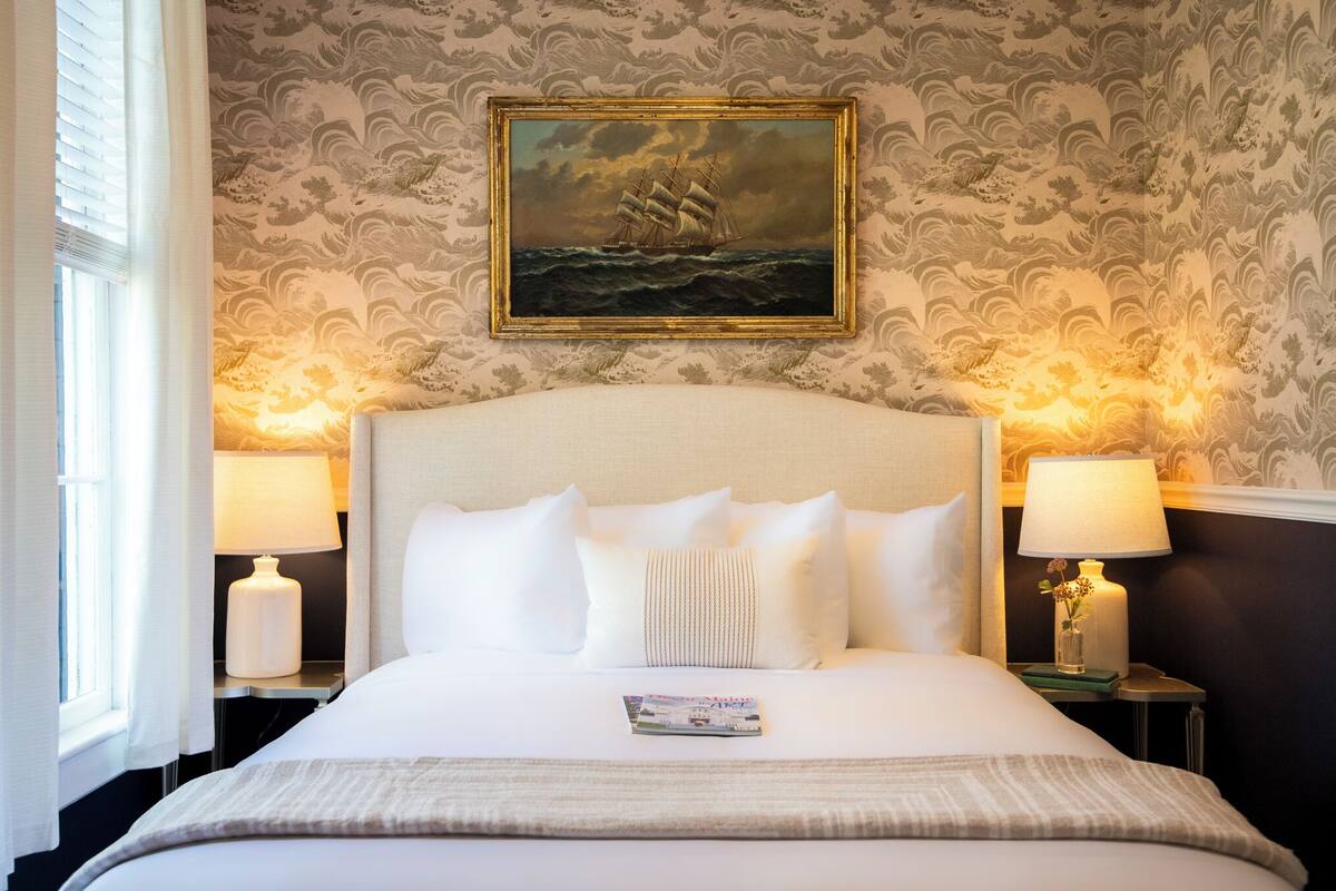 Uma cama bem arrumada com lençóis brancos, um quadro de um navio na parede e duas luminárias de cabeceira.