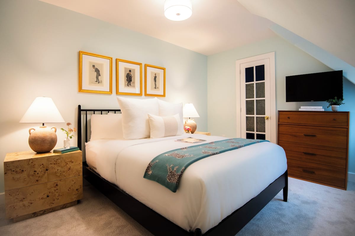 Quarto com cama, duas mesas de cabeceira com luminárias, cômoda, televisão na parede e quadros acima da cama.