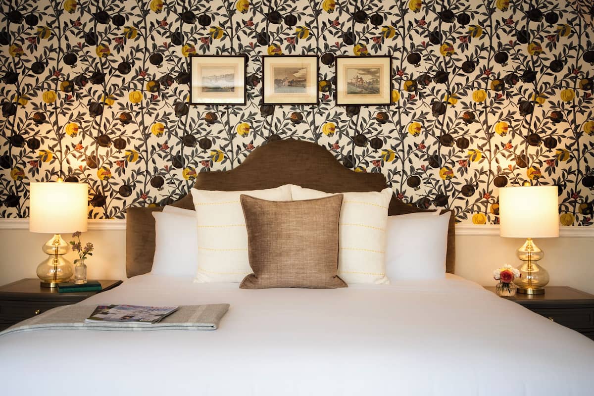 Cama bem arrumada com cabeceira marrom, duas luminárias de apoio e um padrão floral no papel de parede.