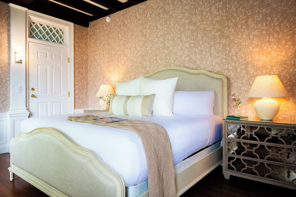 Uma cama bem arrumada com esquema de cores branco e bege, ladeada por duas mesas de cabeceira com abajures, em frente a um papel de parede floral.