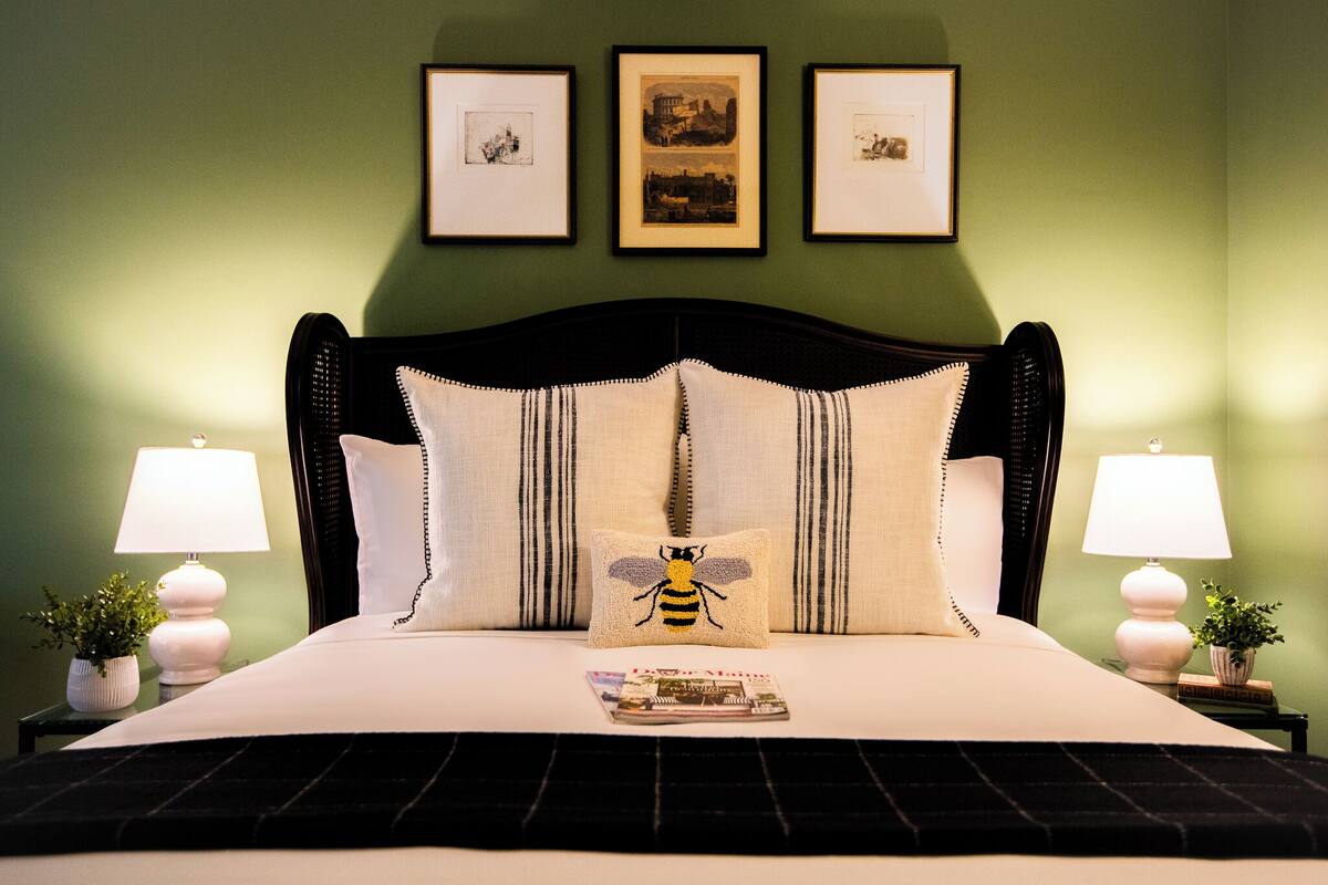 Cama bem arrumada com um travesseiro estampado com um desenho de abelha, acompanhada por duas mesas de cabeceira com luminárias e pequenos vasos de plantas.