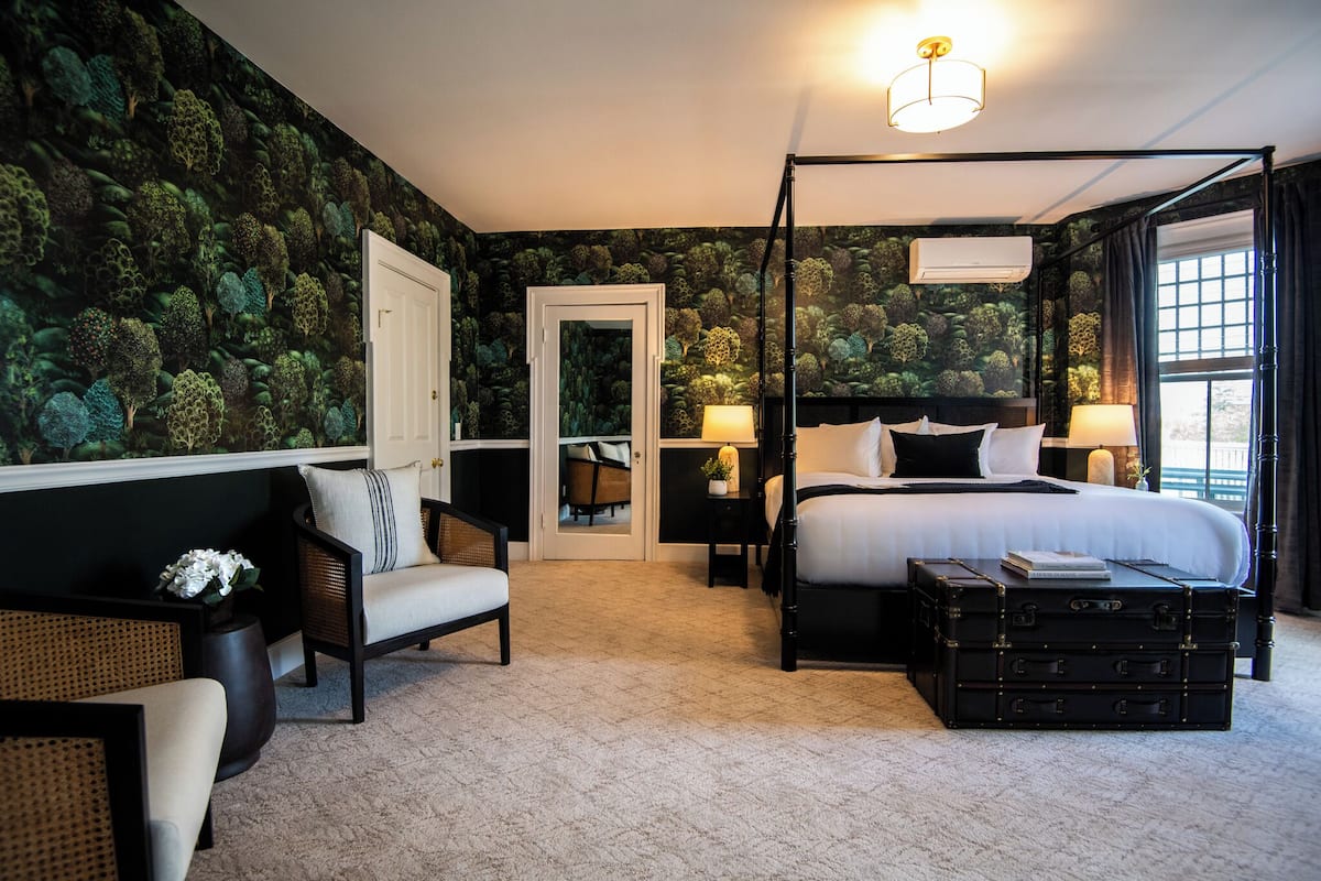Quarto com cama de dossel, um espaço de estar com uma poltrona e uma mesinha, uma janela grande com cortinas, e uma porta que dá para outro cômodo.
