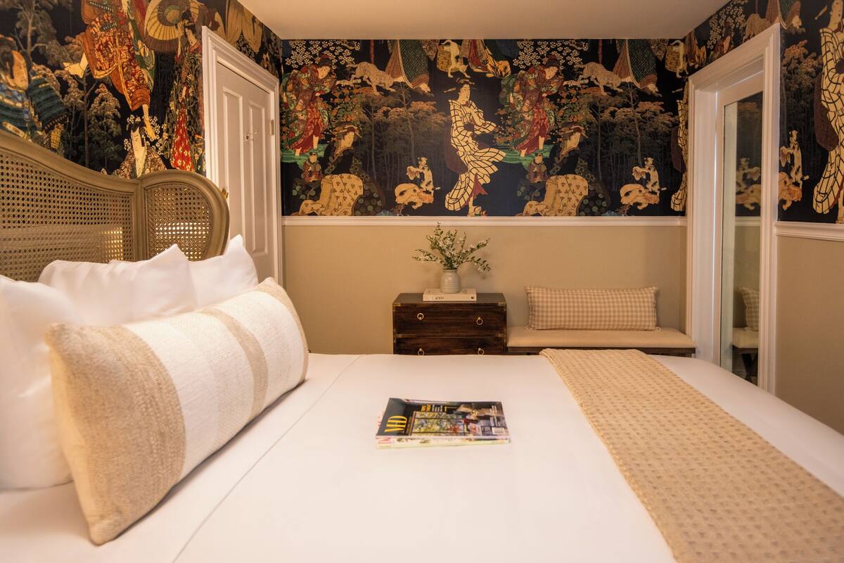 Quarto de hotel com uma cama grande, uma mesa de cabeceira, uma pequena mesa e uma parede com tapeçaria detalhada.