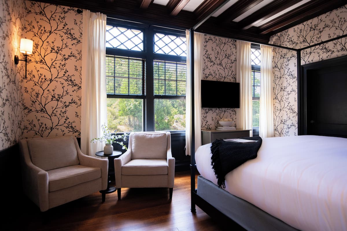Quarto com uma cama grande, duas poltronas, uma mesinha, televisão e janelas amplas com cortinas.