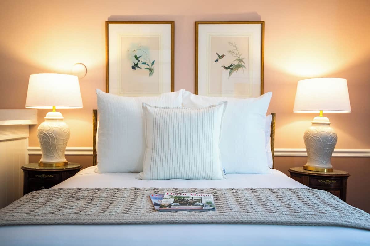 Cama bem arrumada com lençóis brancos e duas almofadas, com duas mesas de cabeceira com luminárias e impressões botânicas emolduradas na parede.