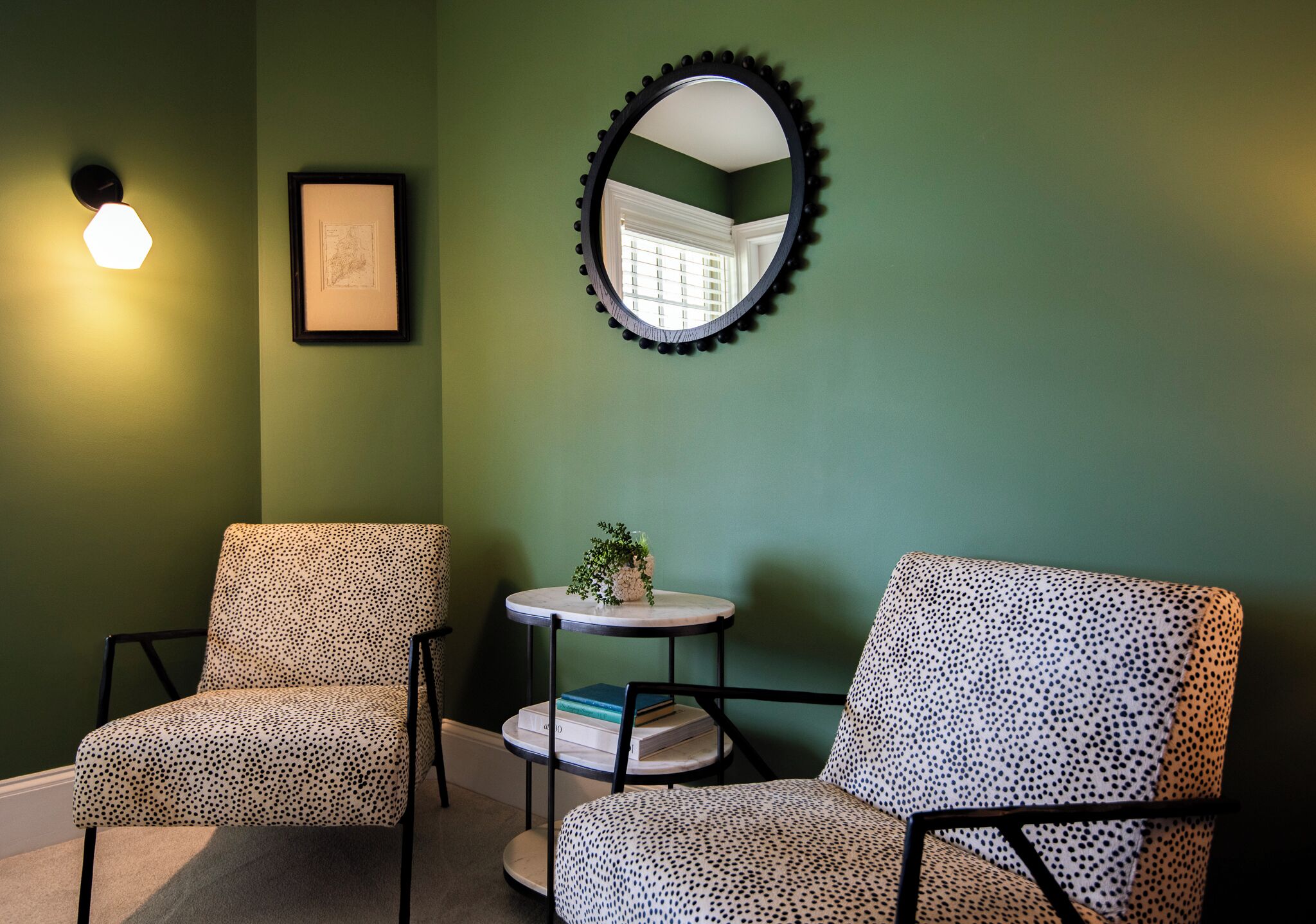 Quarto com duas cadeiras estampadas, uma pequena mesa redonda com uma planta, um quadro emoldurado e um espelho fixado na parede.