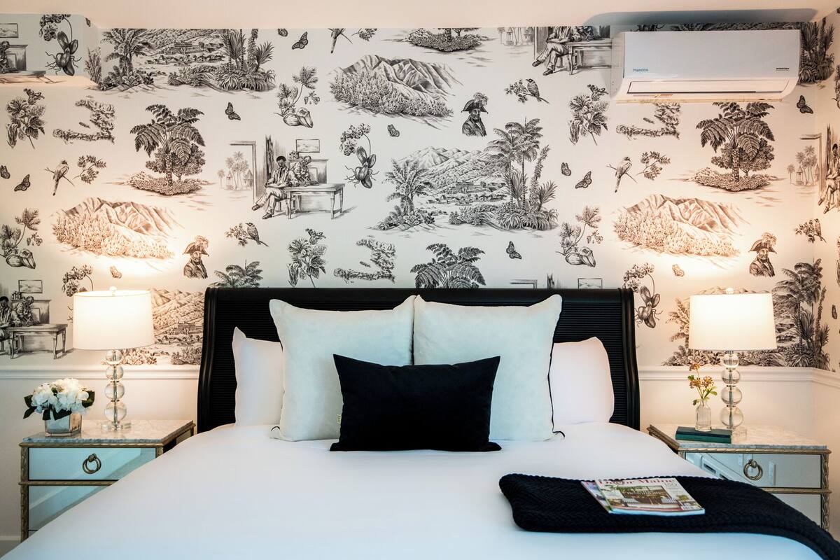 Cama bem arrumada com roupa de cama branca e preta, ladeada por duas mesas de cabeceira com luminárias e objetos decorativos. O quarto apresenta uma cabeceira grande e escura e uma parede com um mural detalhado em preto e branco.