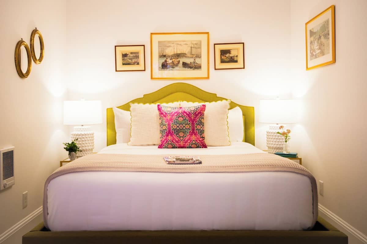 Uma cama bem arrumada com cabeceira verde, duas luminárias de mesa e quadros nas paredes.