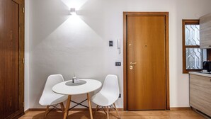 Quarto pequeno e moderno, com porta de madeira, mesa redonda com duas cadeiras brancas, luminária na parede e janela com padrão geométrico.
