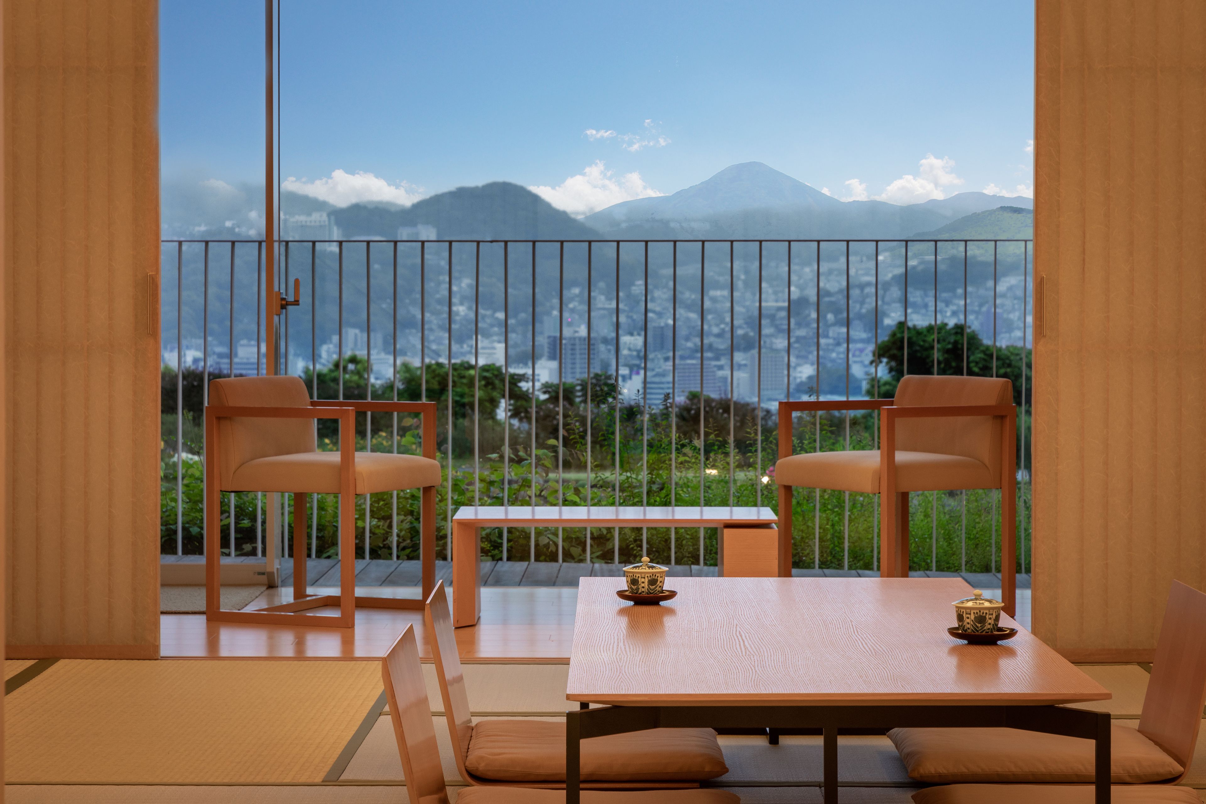 Uma varanda com mesa e cadeiras de madeira, vista para montanhas e paisagem urbana ao fundo.