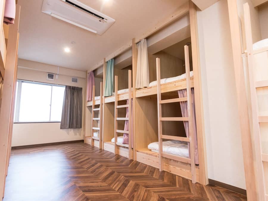 Quarto com beliches, piso de madeira em padrão herringbone, janela com cortinas e aparelho de ar condicionado.