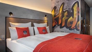 Quarto de hotel moderno com uma cama grande, duas almofadas com etiquetas, um cobertor vermelho e uma parede com grafite.