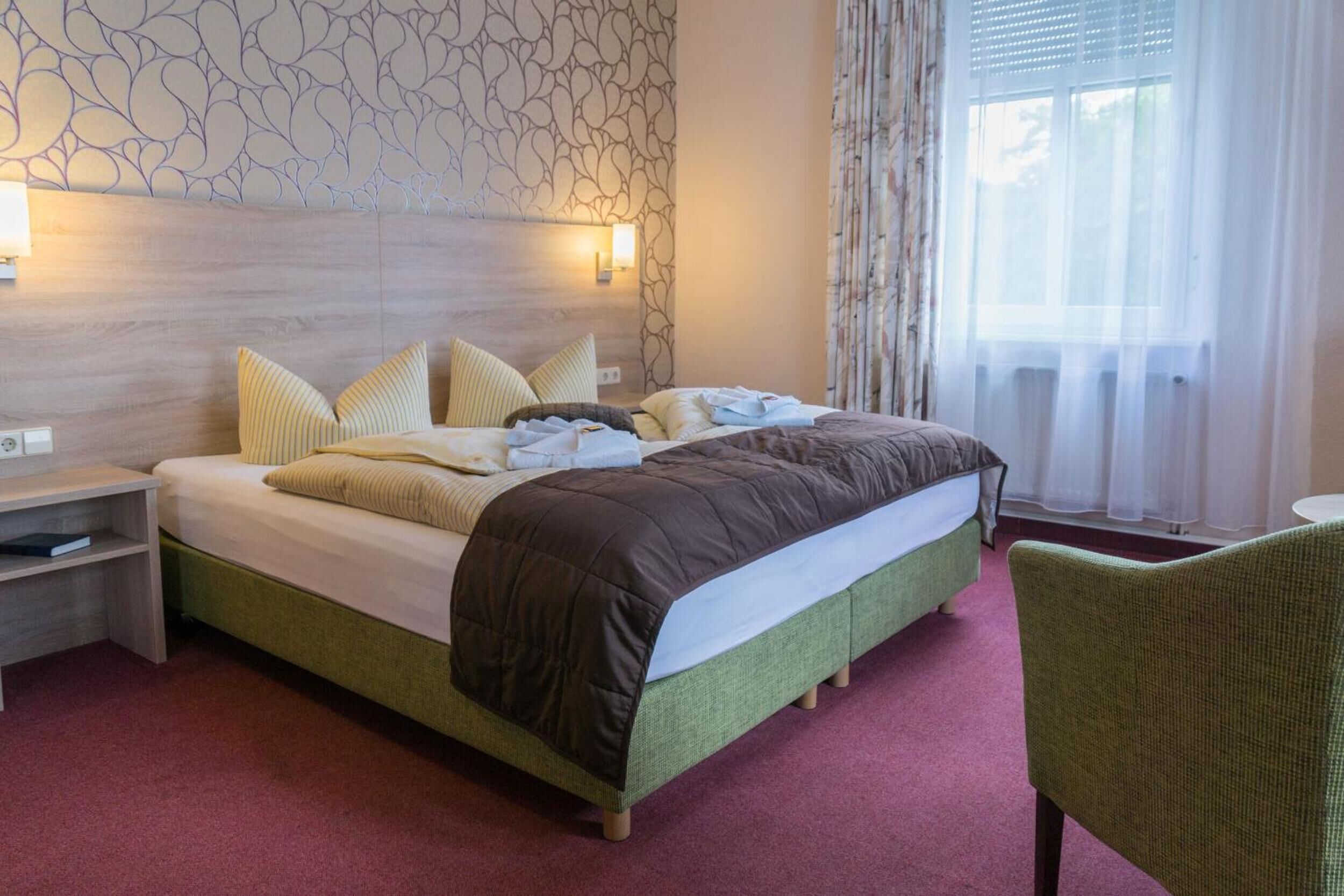 Quarto de hotel com cama, mesa de cabeceira, cadeira e janela com cortinas.