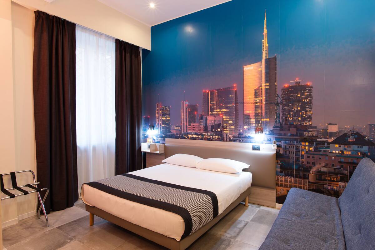 Quarto de hotel com cama, vista para a cidade, janela com cortinas e mesa de cabeceira.