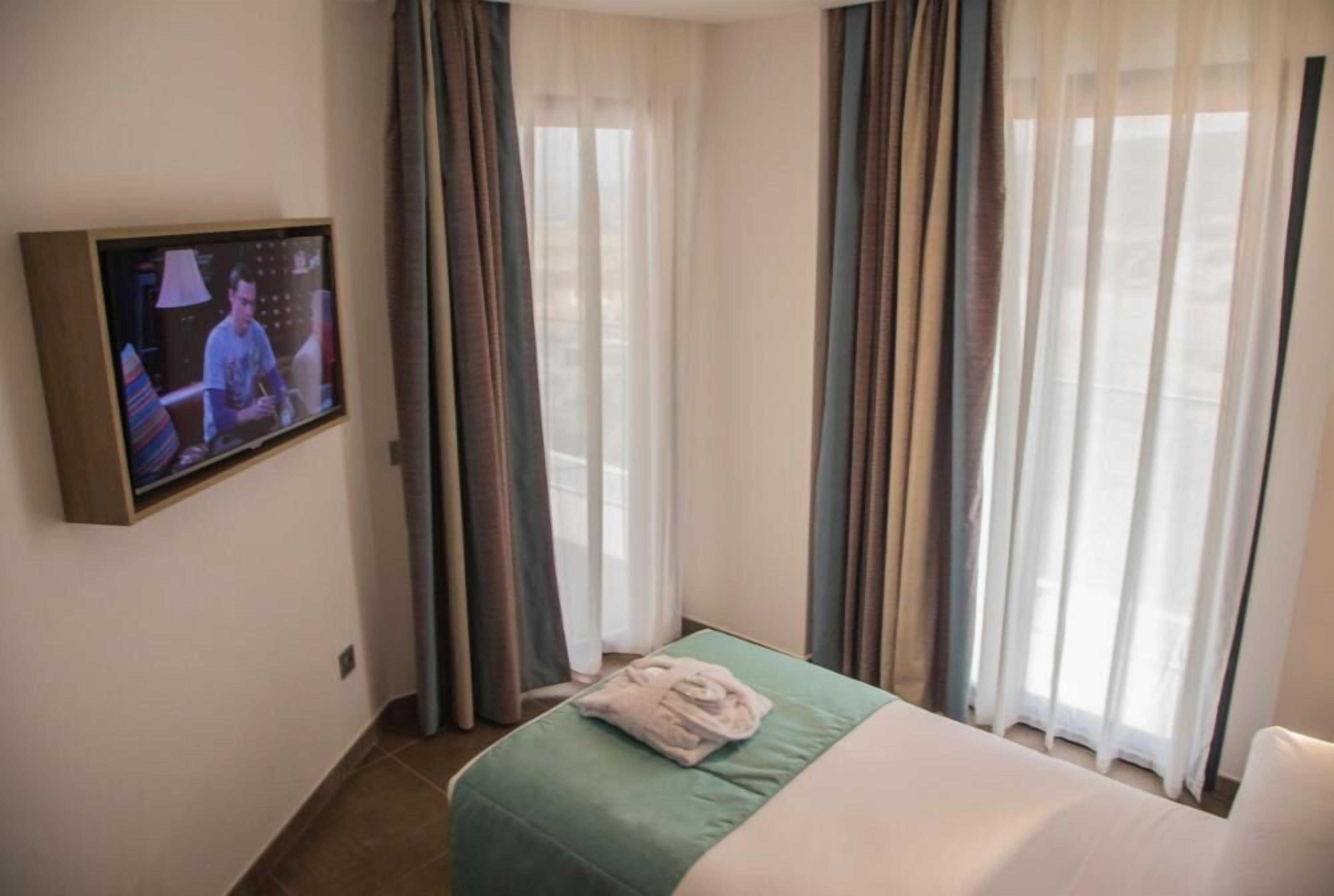Quarto de hotel com cama, televisão na parede e cortinas nas janelas.
