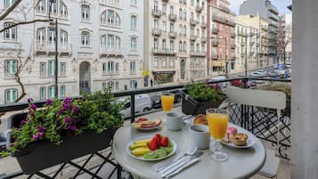 Varanda com mesa posta para o café da manhã, incluindo frutas, doces e bebidas, com vista para uma rua da cidade com edifícios históricos.