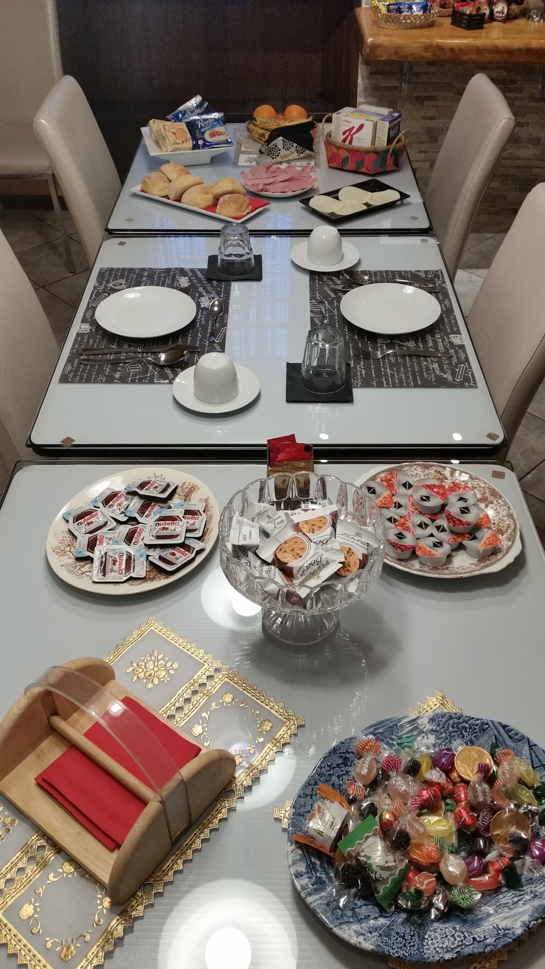 Mesa de jantar posta com pratos, talheres e diversos alimentos, incluindo doces e chocolates, num quarto de hotel.