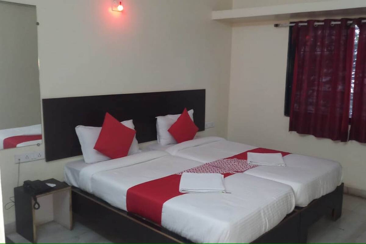 Uma cama de casal com roupa de cama branca e detalhes vermelhos, uma mesa de cabeceira, uma luminária fixada na parede e uma cortina vermelha.