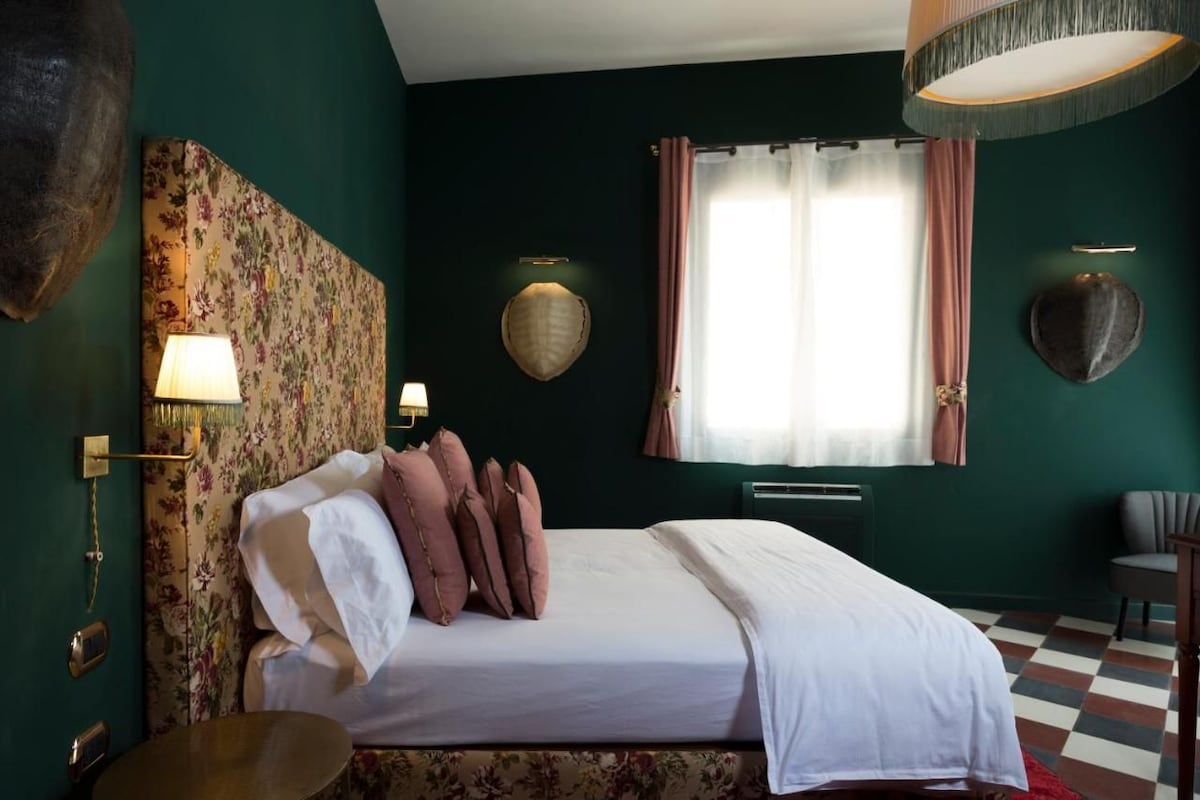 Quarto com cama, mesa de cabeceira, luminária, relógio na parede e janela com cortinas.