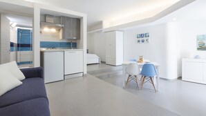 Uma sala de estar moderna e bem iluminada, com sofá, mesa de jantar com cadeiras, cozinha compacta e uma cama.