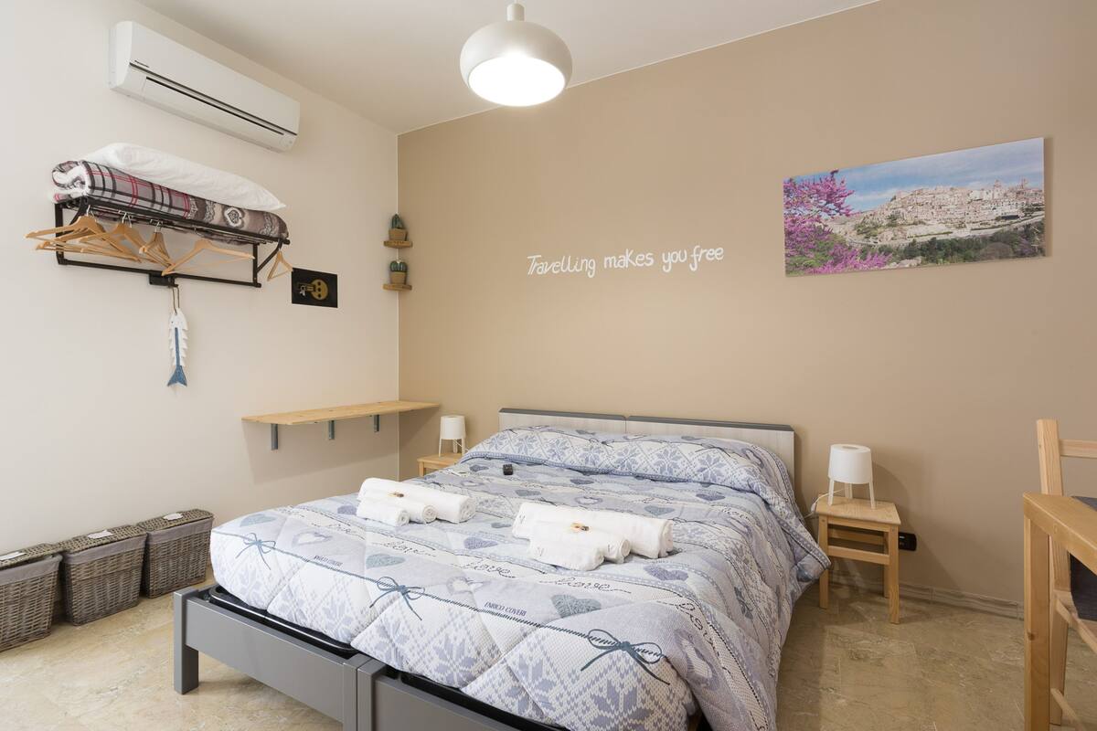 Quarto bem arrumado com cama, mesas de cabeceira, ar condicionado fixado na parede e uma frase motivacional.