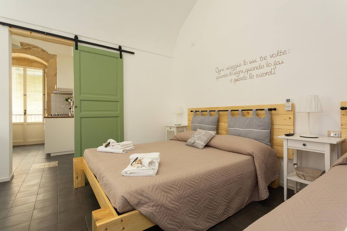 Quarto com porta deslizante verde, cama com colcha bege, duas mesas de cabeceira com abajures e uma parede com uma citação em italiano.