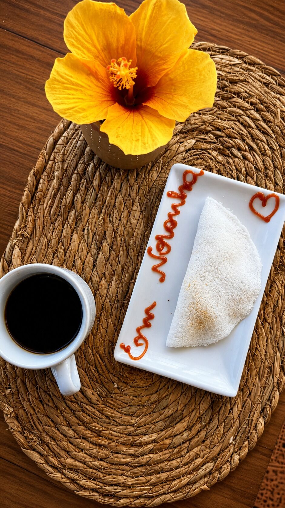 Um jogo americano tecido com um prato de arroz branco, uma chávena de café preto e uma flor amarela.