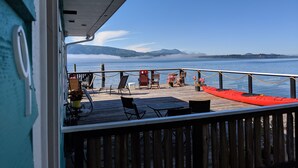 Um deck de madeira com cadeiras e um caiaque vermelho, com vista para um lago calmo e montanhas ao longe.