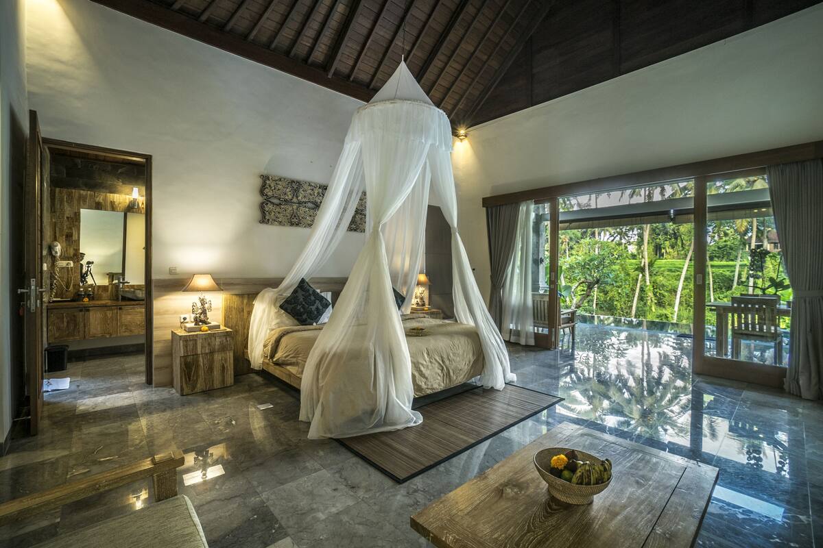 Quarto com cama de dossel, mesa de cabeceira de madeira, espelho e vista para o exterior.