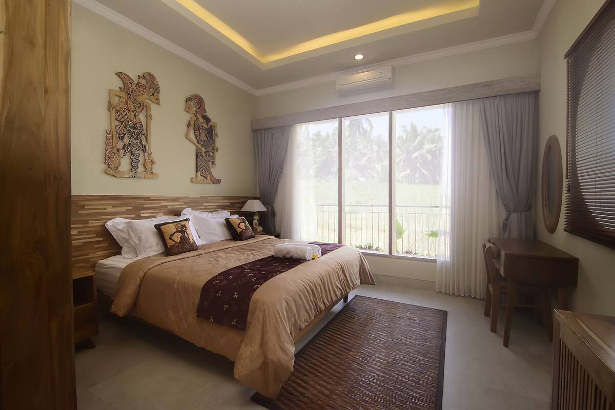 Quarto bem iluminado com uma cama grande, mesa de cabeceira de madeira, escrivaninha e janela com cortinas.