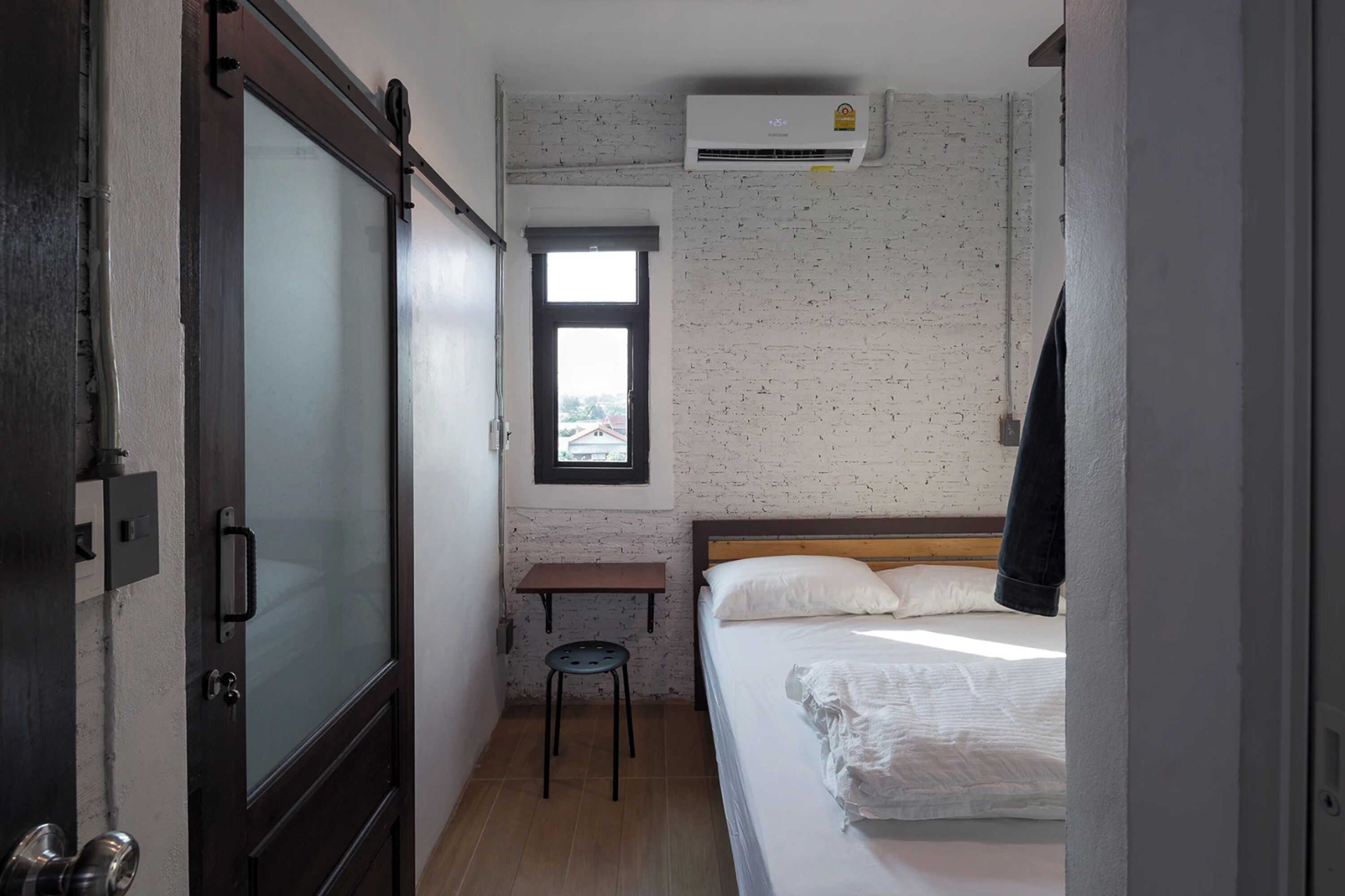 Quarto com cama, um banquinho, janela e ar-condicionado.