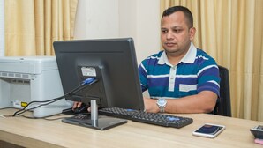Uma pessoa sentada em uma mesa com um computador, uma impressora e um smartphone.