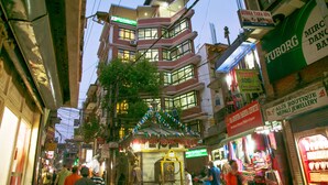 Uma rua estreita com edifícios altos, lojas e um pequeno santuário com decorações coloridas.