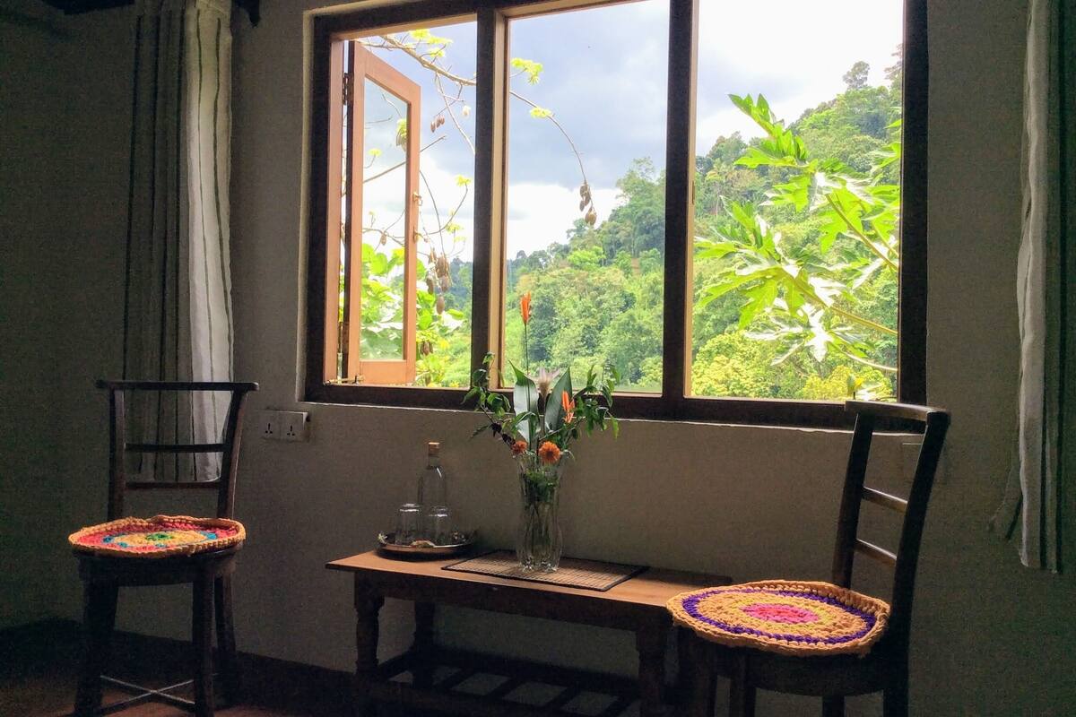 Quarto com uma grande janela, duas cadeiras com assentos trançados, uma pequena mesa com um vaso de flores e vista para vegetação exuberante.