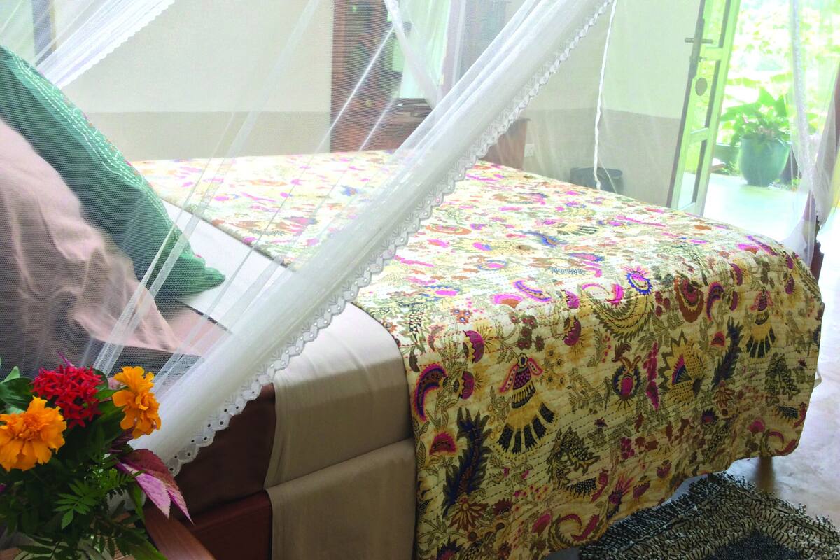 Uma cama com colcha estampada em flores coloridas, um dossel branco e vista para o verde lá fora.