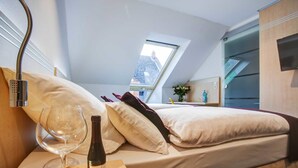 Quarto com cama, mesa de cabeceira, garrafa de vinho e copo, janela e claraboia.