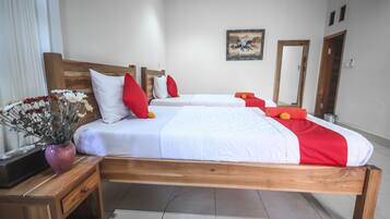 Quarto de hotel com duas camas, cabeceiras de madeira, roupa de cama branca, almofadas vermelhas e brancas, um vaso com flores e um quadro na parede.
