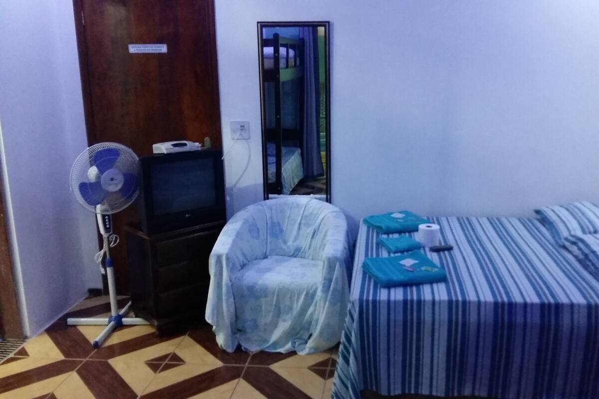 Quarto com cama, cadeira, televisão e ventilador.