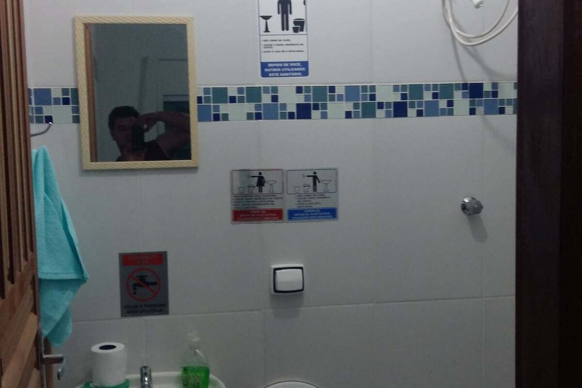 Um banheiro pequeno com vaso sanitário, pia e espelho. Há placas na parede, secador de mãos e área de chuveiro.