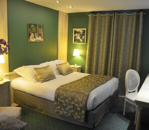 Quarto de hotel com cama, escrivaninha, cadeira, luminária e duas pinturas na parede.