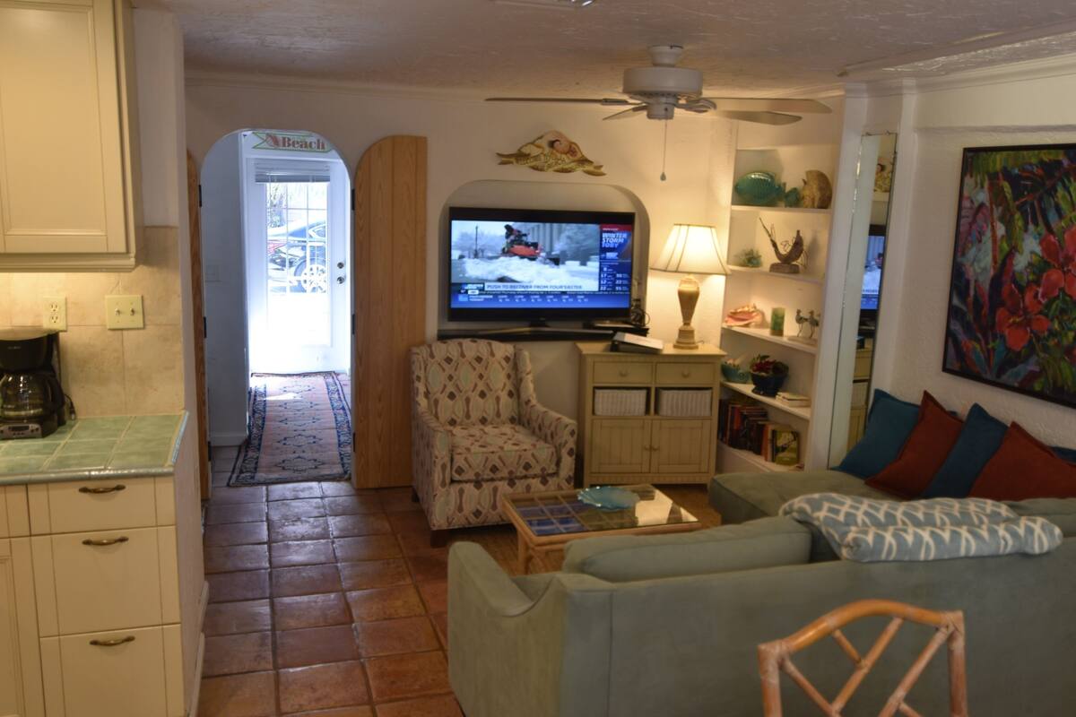 Uma sala de estar com televisão, uma poltrona estampada, um sofá e um ventilador de teto.