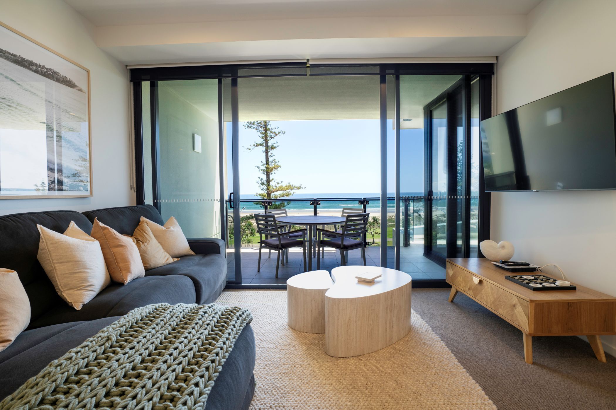 Uma sala de estar moderna com sofá, mesa de centro e televisão, com vista para uma varanda e o oceano.