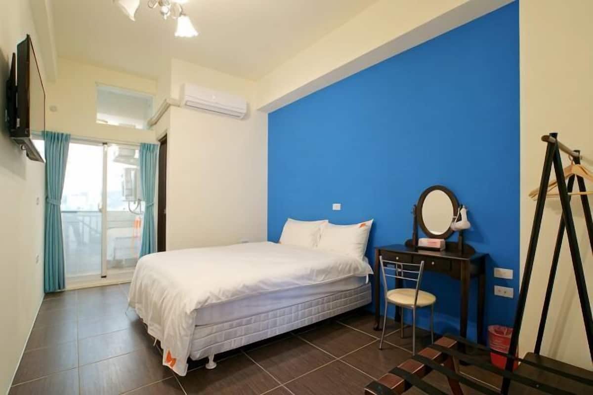 Quarto com cama, escrivaninha com espelho, cadeira, janela com cortinas e porta que dá para uma varanda.