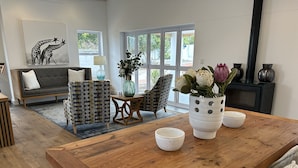 Uma sala de estar moderna com uma mesa de centro de madeira, duas poltronas estampadas, um sofá e uma janela grande com vista para o verde.